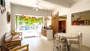 Uma sala de estar ampla com uma mesa de jantar e cadeiras, um sofá, um ventilador de teto e vista para um jardim através de uma porta de vidro.
