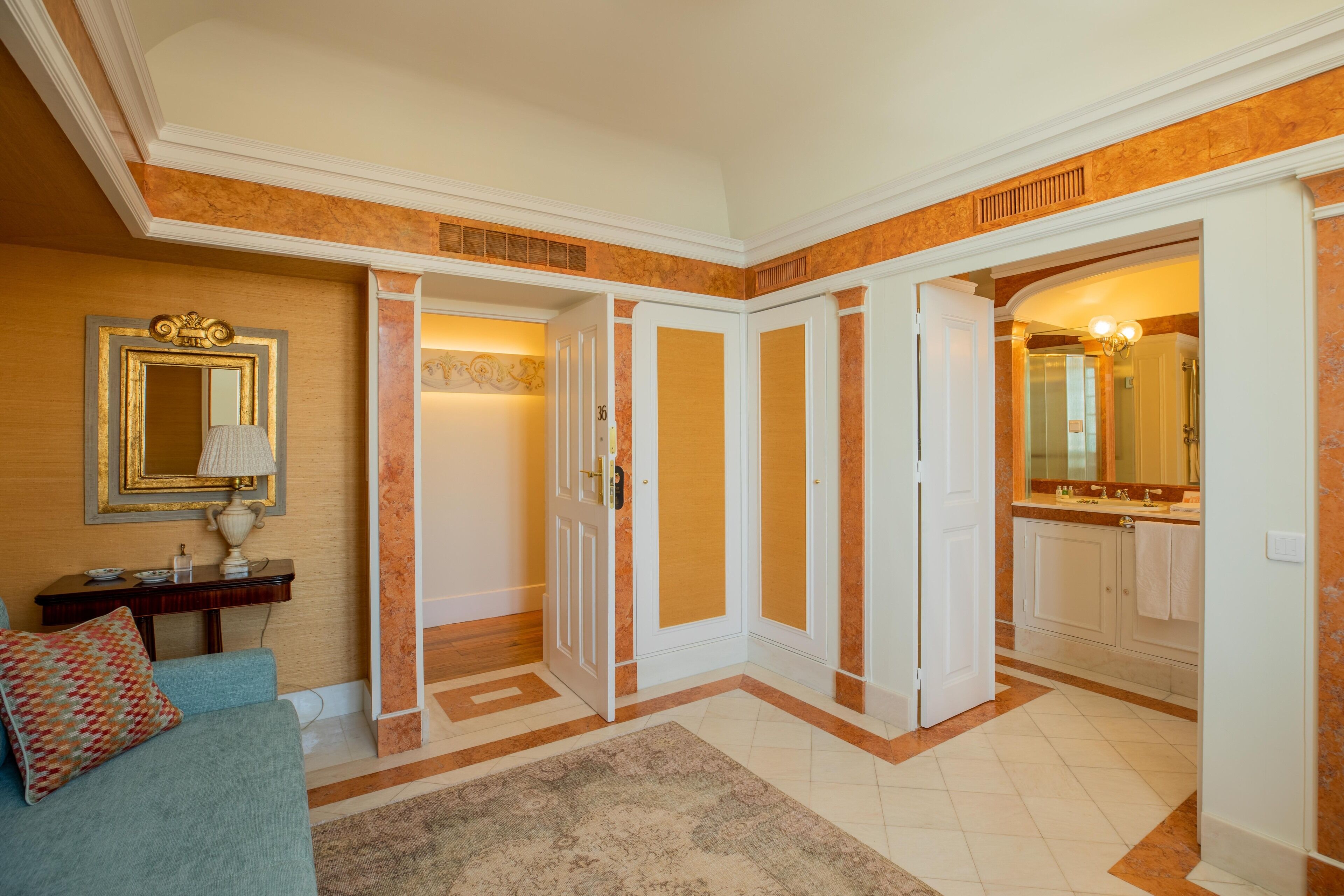 Quarto de hotel com sofá, espelho, porta para um banheiro e piso ladrilhado.