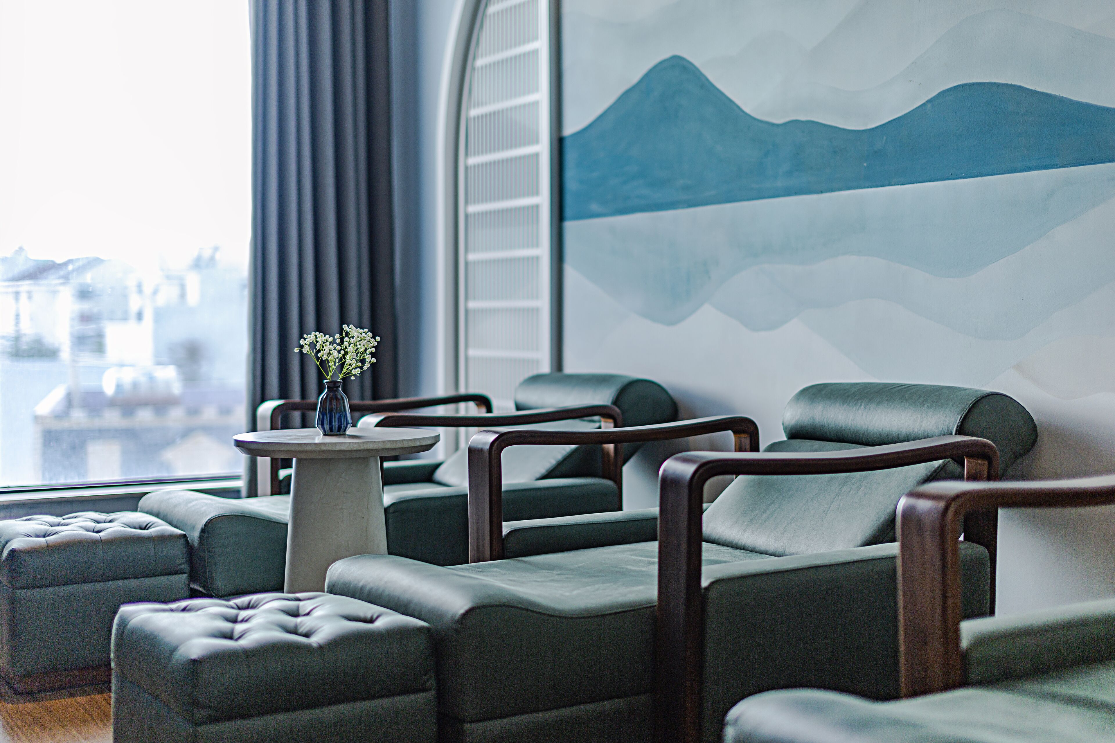 Quarto de hotel moderno com assentos estofados em verde, uma mesa redonda pequena com um vaso e uma janela grande com vista para prédios.