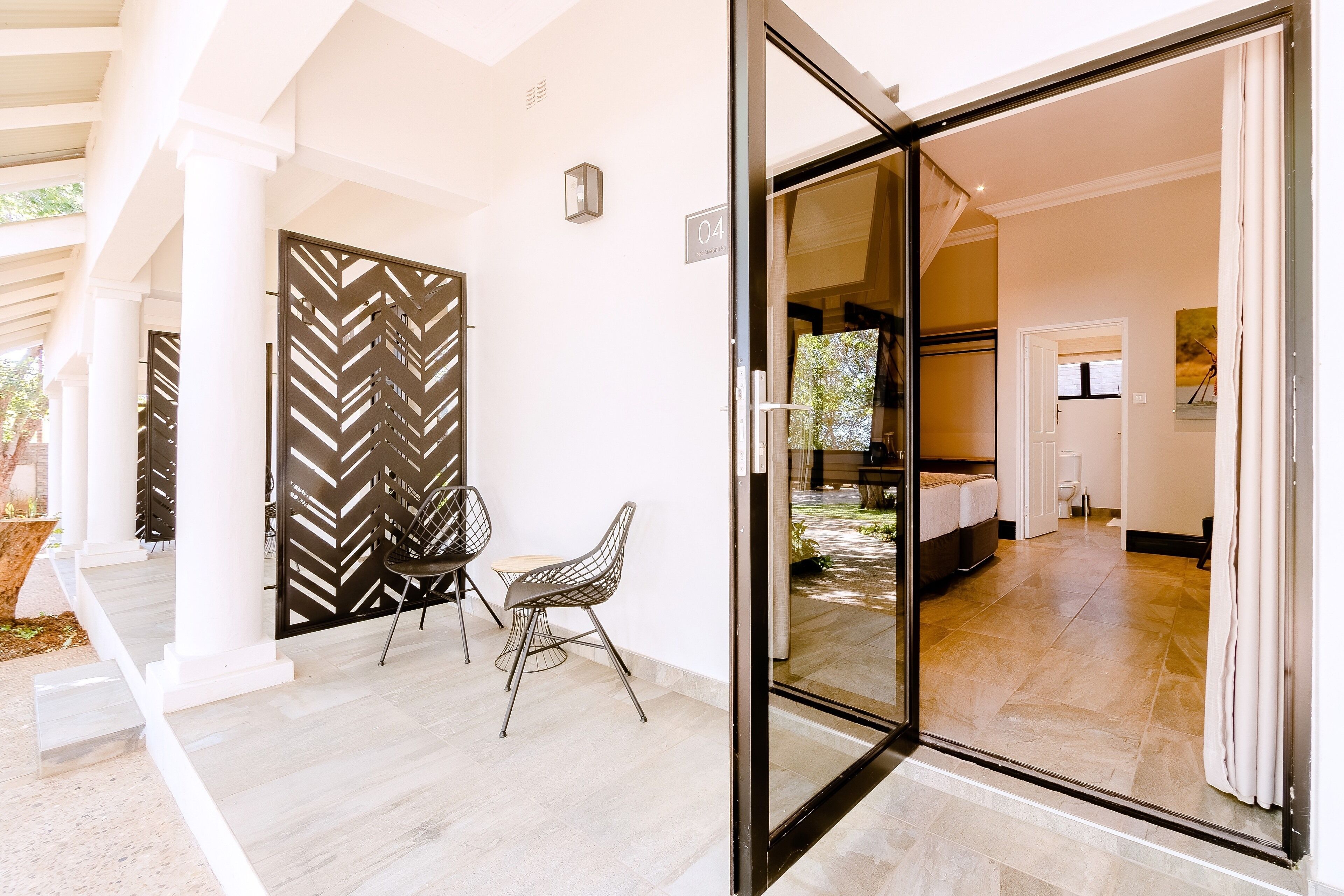 Interior moderno de hotel com porta de vidro que dá para um quarto, área de estar com cadeiras e painel decorativo na parede.