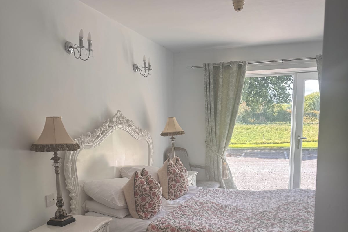 Quarto com cama, duas mesas de cabeceira, luminária, luminária fixa na parede e vista para um jardim pela janela.