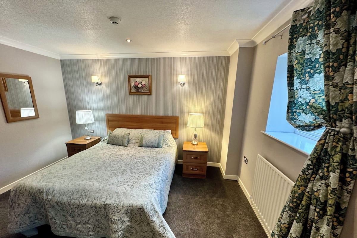 Quarto com cama, duas mesas de cabeceira com abajures, um espelho, janela com cortinas e radiador.