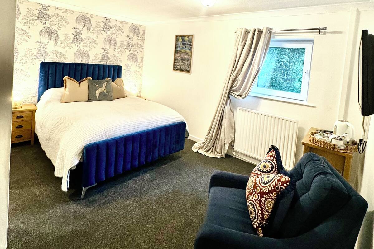 Quarto com cama, mesa de cabeceira, janela com cortinas, radiador e sofá com almofada estampada.