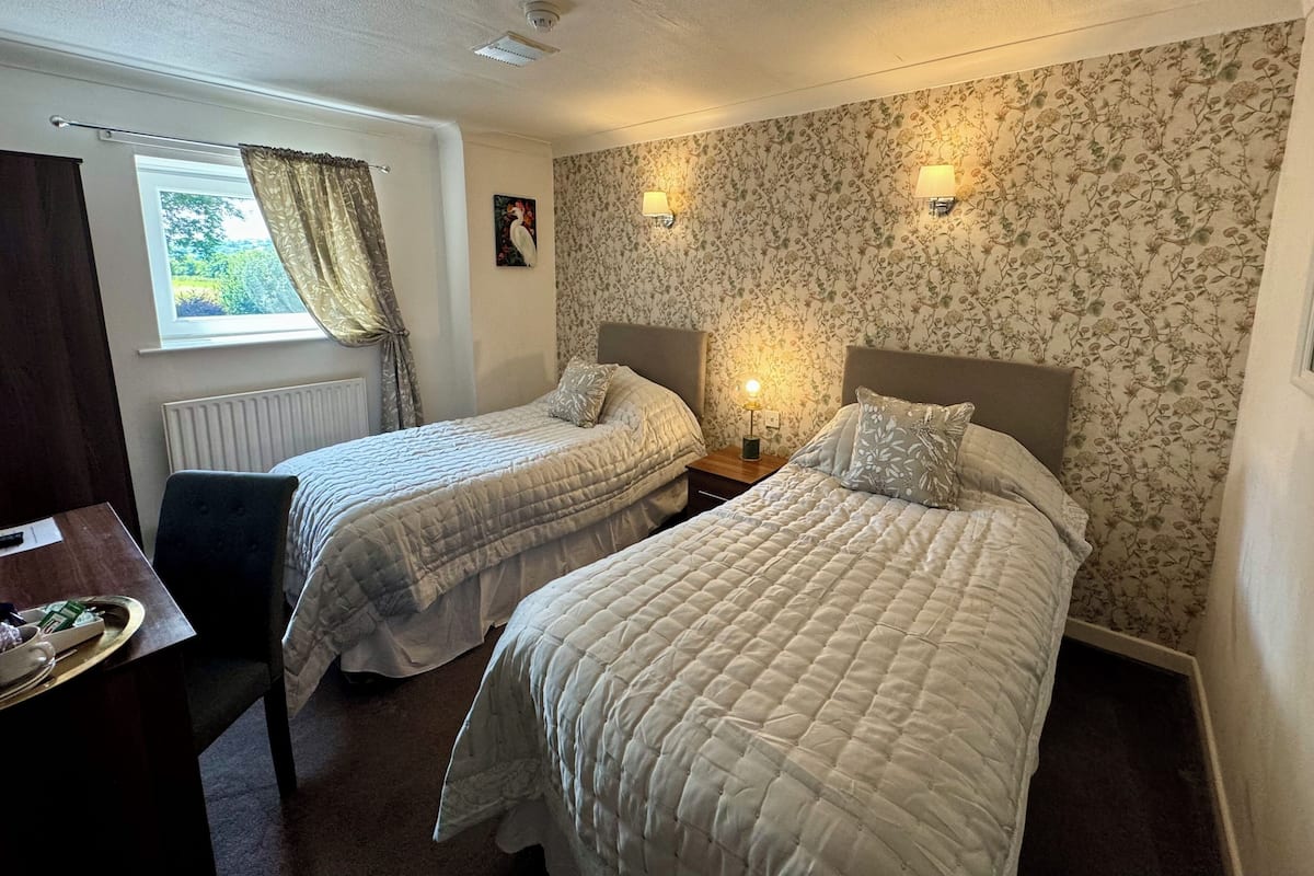 Quarto com duas camas, uma mesinha com bandeja, uma cadeira, janela com cortinas, e parede com papel de parede floral e luminárias.