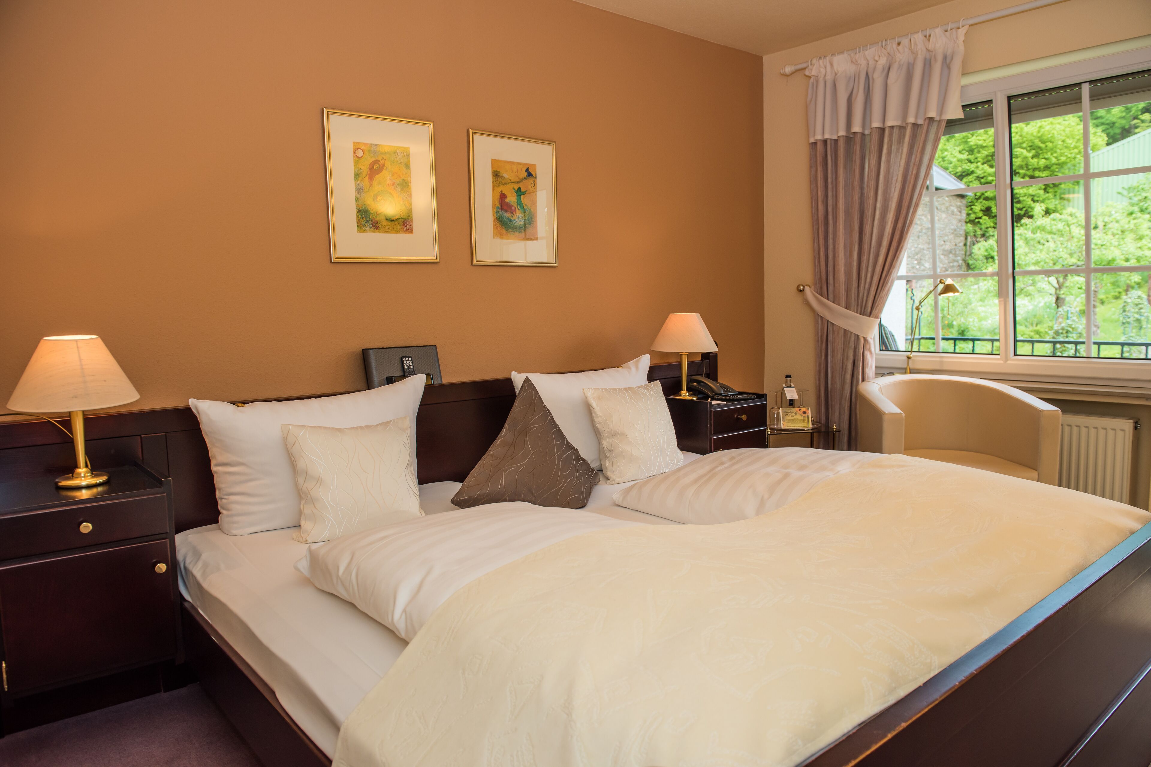 Quarto de hotel com uma cama grande, duas mesas de cabeceira com luminárias, uma cadeira e duas obras de arte emolduradas na parede.