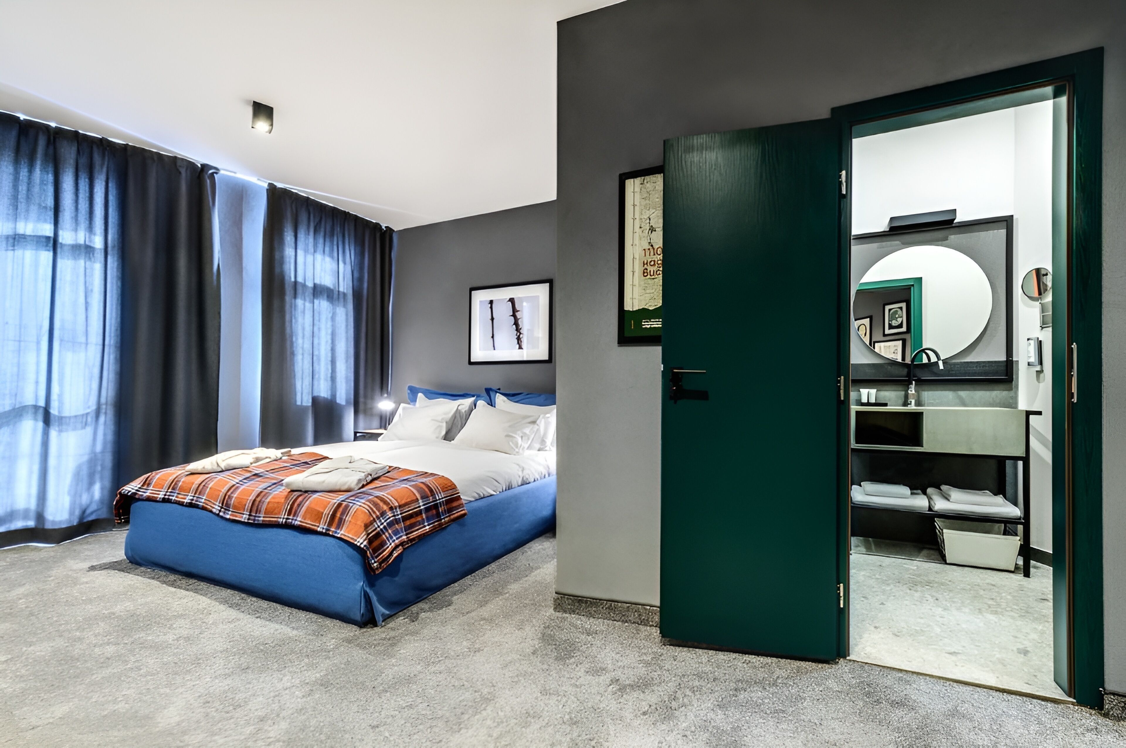 Quarto moderno com cama, cortinas, porta para um banheiro e espelho fixado na parede.