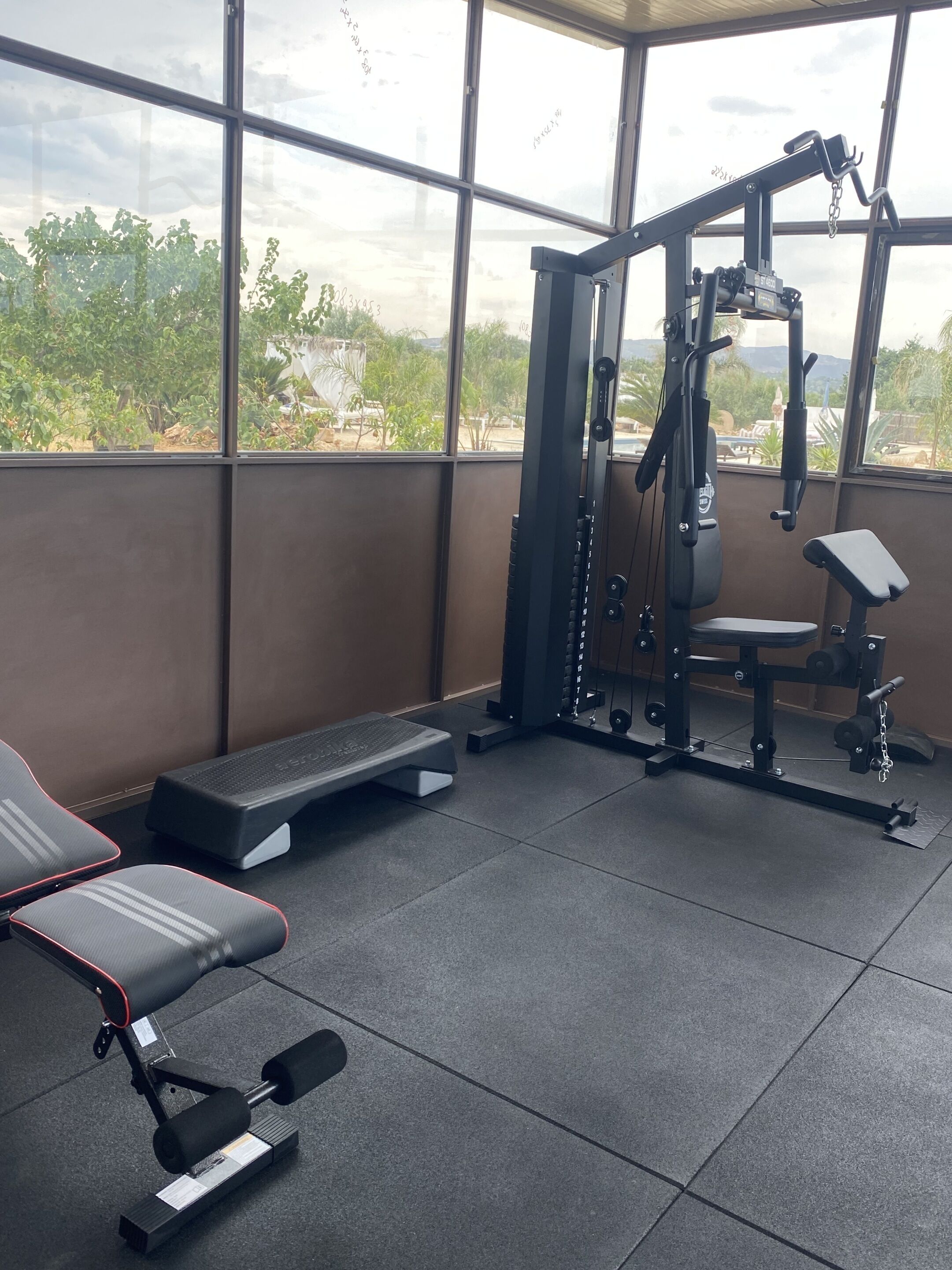 Uma academia com equipamentos de ginástica, um banco e vista para o verde através de amplas janelas.