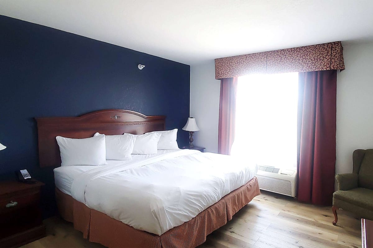 Quarto de hotel com uma cama grande, mesa de cabeceira, abajur, cadeira e janela com cortinas.