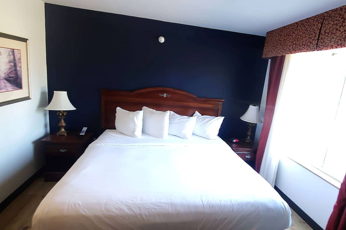 Quarto de hotel com uma cama grande, duas mesas de cabeceira, um criado-mudo, um relógio de parede e uma janela com cortinas.