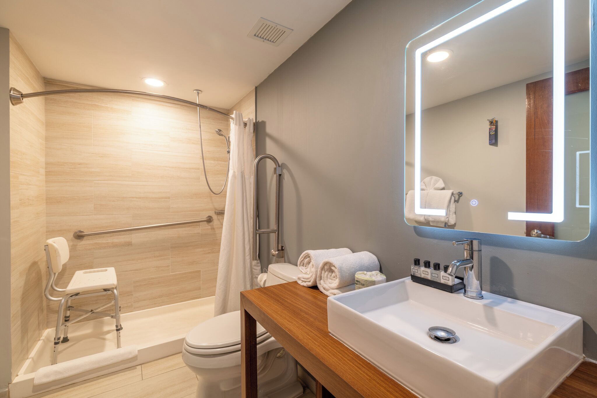 Uma casa de banho moderna com chuveiro, sanita e lavatório, equipada com um apoio de proteção e um espelho.