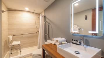 Uma casa de banho moderna com chuveiro, sanita e lavatório, equipada com um apoio de proteção e um espelho.