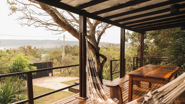 Um deck de madeira com uma rede, uma mesa e um banco, com vista para um jardim e colinas ao longe.