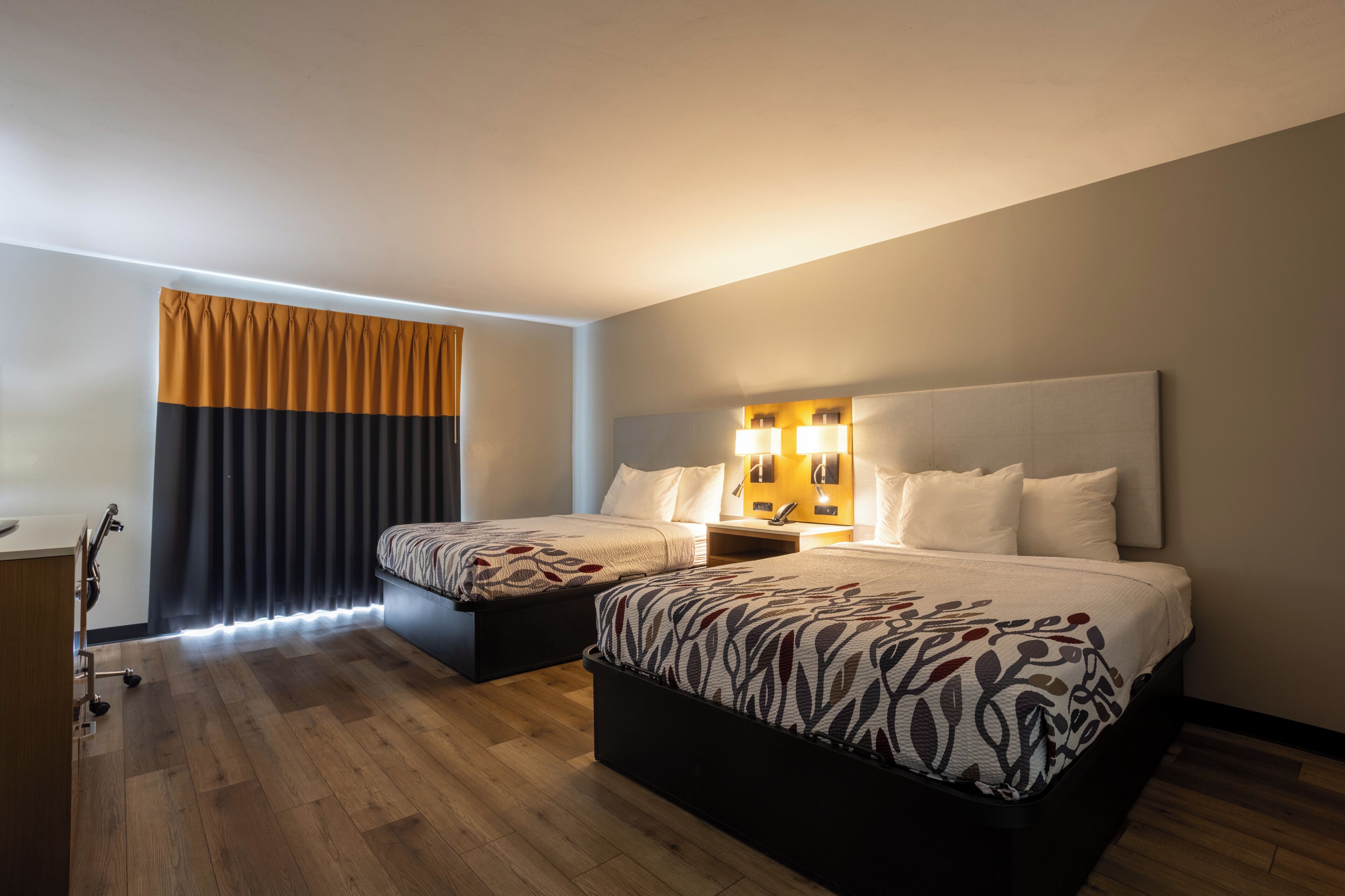 Quarto de hotel com duas camas, uma mesinha de cabeceira, uma luminária e uma janela com cortinas.