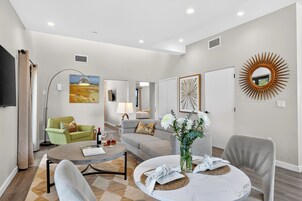 Uma sala de estar moderna com sofá, poltronas, mesa de centro e mesa de jantar. Há um quadro na parede, um abajur e um espelho decorativo.