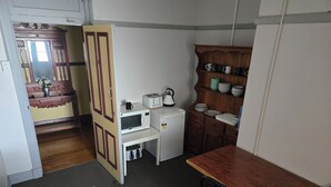 Uma cozinha compacta com micro-ondas, torradeira e chaleira sobre um balcão branco. Há uma mesa de madeira e uma cadeira, e um armário com pratos e copos.