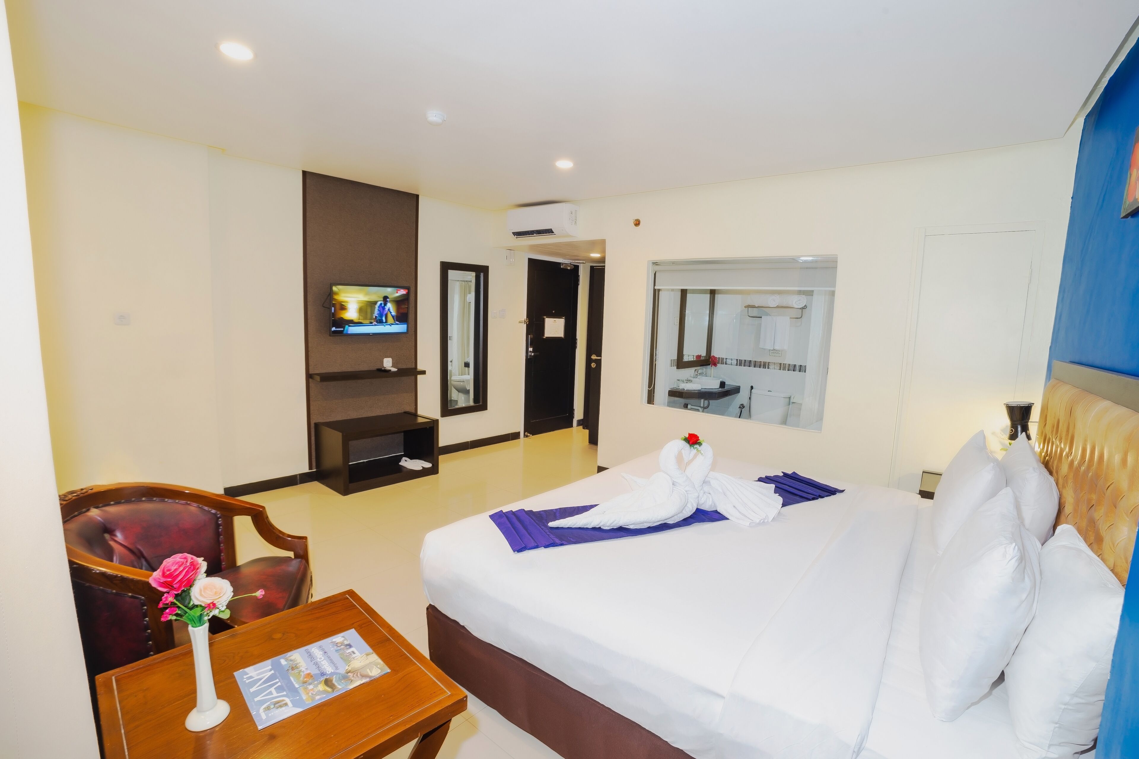 Quarto de hotel com uma cama grande, televisão, escrivaninha com um vaso de flores e banheiro visível através de uma porta de vidro.