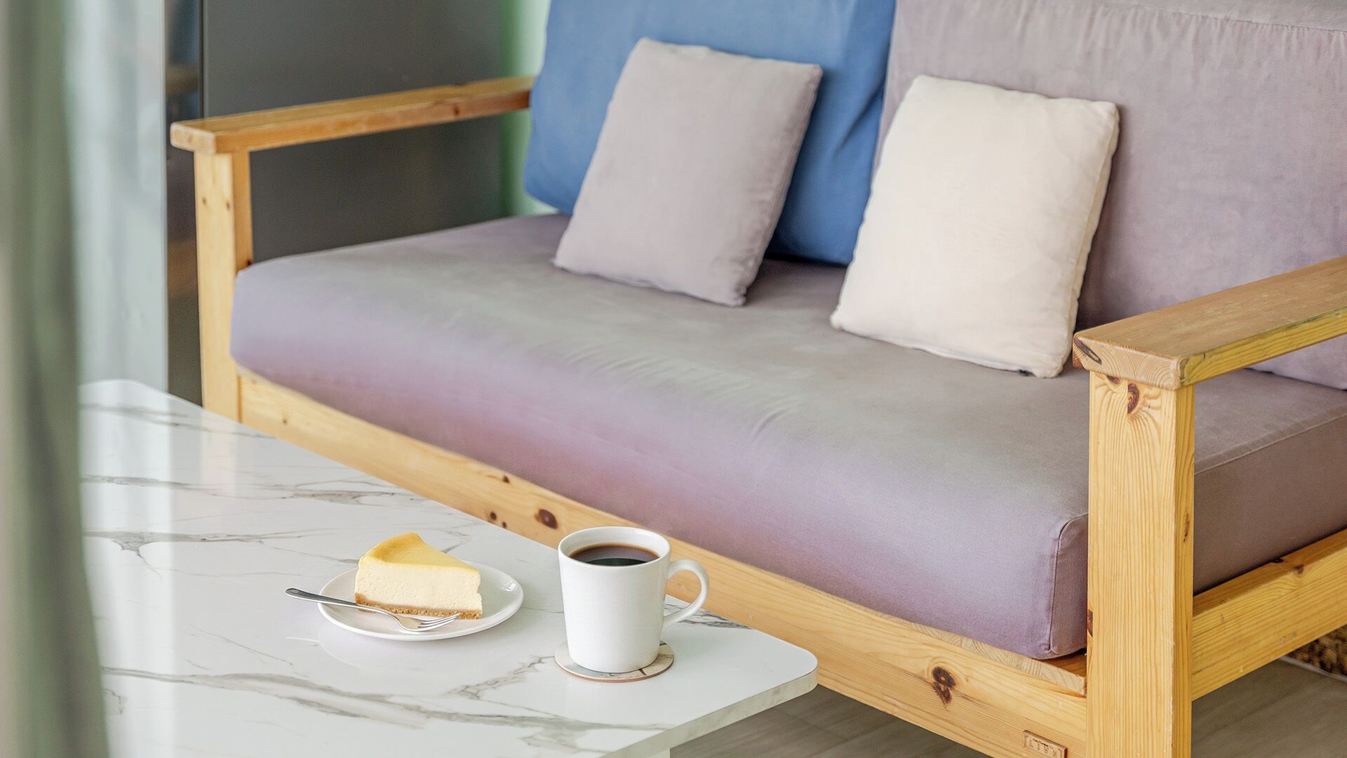 A wooden-framed sofa with cushions, a marble table with a slice of cheesecake and a cup of coffee, and a green wall in the background.