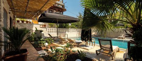 Uma área à beira da piscina com espreguiçadeiras, piscina e um espaço de estar coberto com telhado de palha.