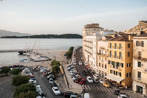Uma rua costeira com carros estacionados, uma marina com veleiros e edifícios com varandas.