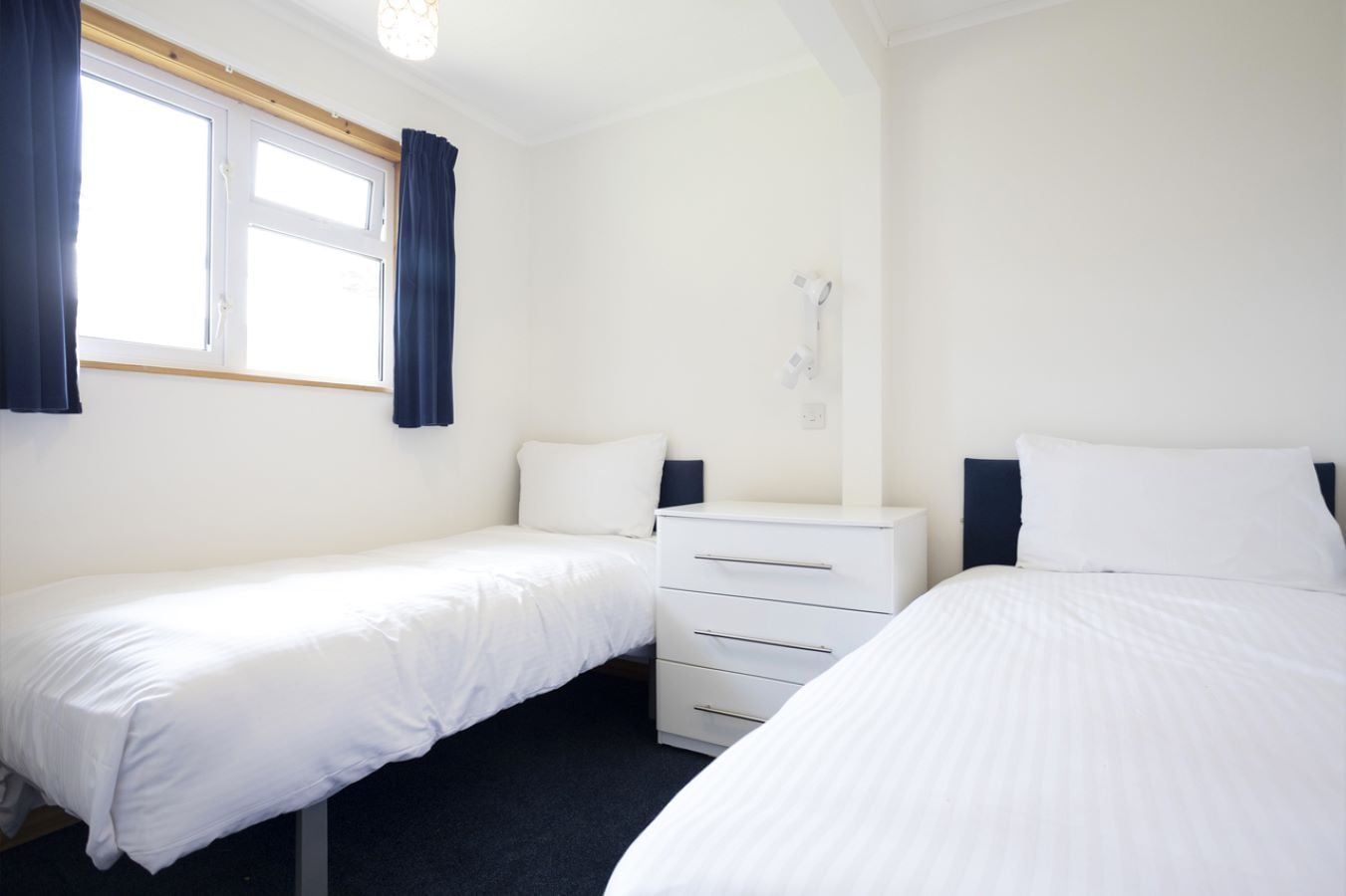 Quarto com duas camas de solteiro, cômoda branca e janela com cortinas.
