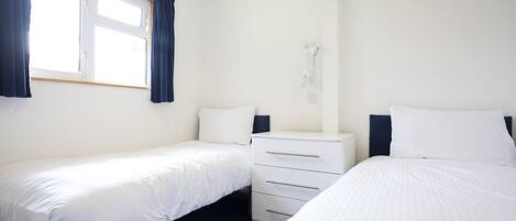 Quarto com duas camas de solteiro, cômoda branca e janela com cortinas.