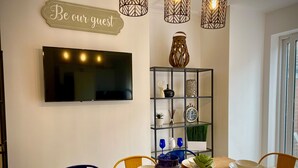 Uma área de refeições com mesa de madeira posta para quatro pessoas, uma TV na parede e um letreiro com a mensagem 'Seja nosso convidado'.