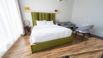 Quarto de hotel moderno com uma cama grande, um sofá cinza, uma poltrona cinza e piso de madeira.