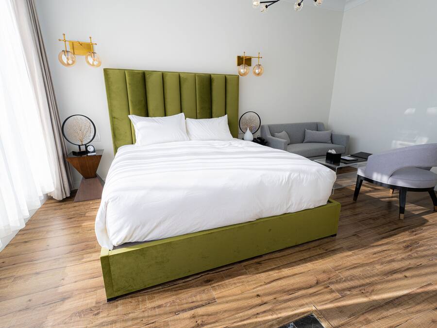 Quarto de hotel moderno com uma cama grande, um sofá cinza, uma poltrona cinza e piso de madeira.