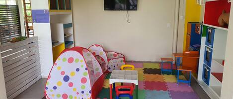 Uma sala de jogos infantil com tendas coloridas, uma mesa pequena e uma TV fixada na parede.
