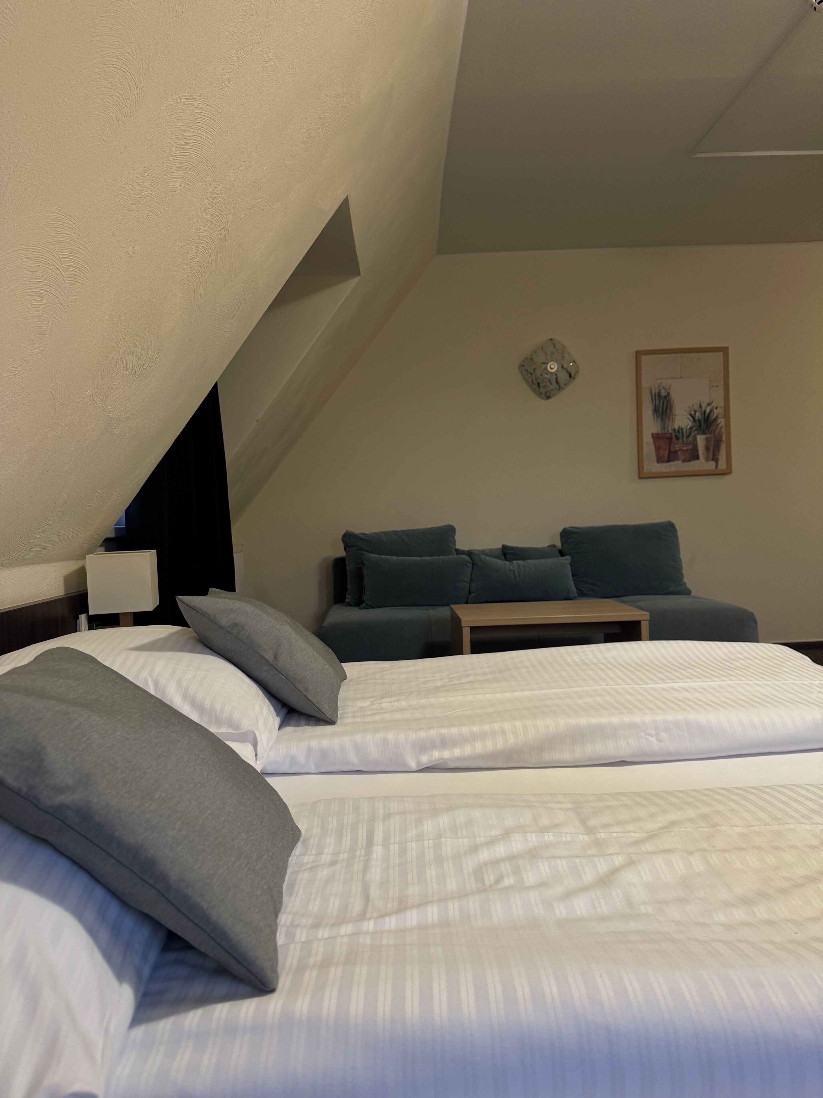 Quarto com teto inclinado, cama com roupa de cama branca, mesa de cabeceira e um espaço de estar com almofadas.