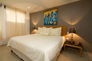 Quarto bem iluminado com uma cama grande, duas luminárias de cabeceira, um quadro na parede e uma mesa de cabeceira com um livro.