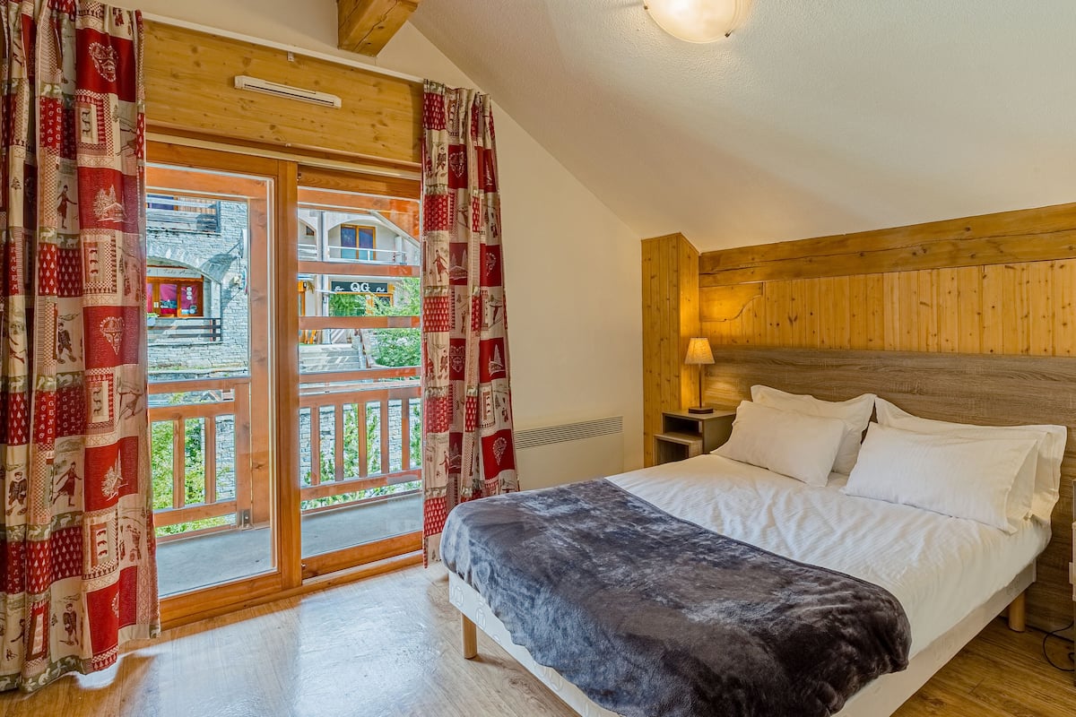 Quarto com cabeceira de madeira, cama com roupa de cama branca, luminária de cabeceira e vista para uma varanda com cortinas estampadas.