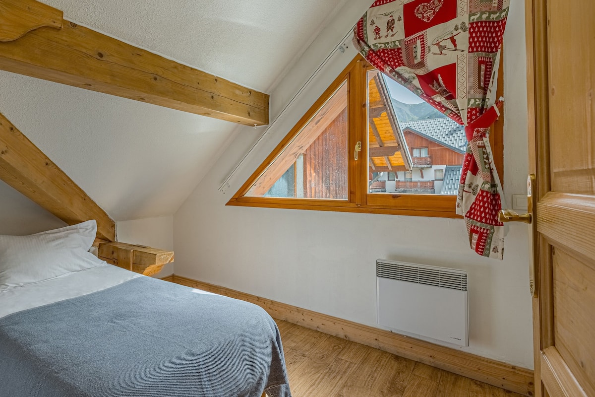 Quarto com teto inclinado, uma cama, uma mesinha de cabeceira, uma janela com vista para edifícios e um radiador.