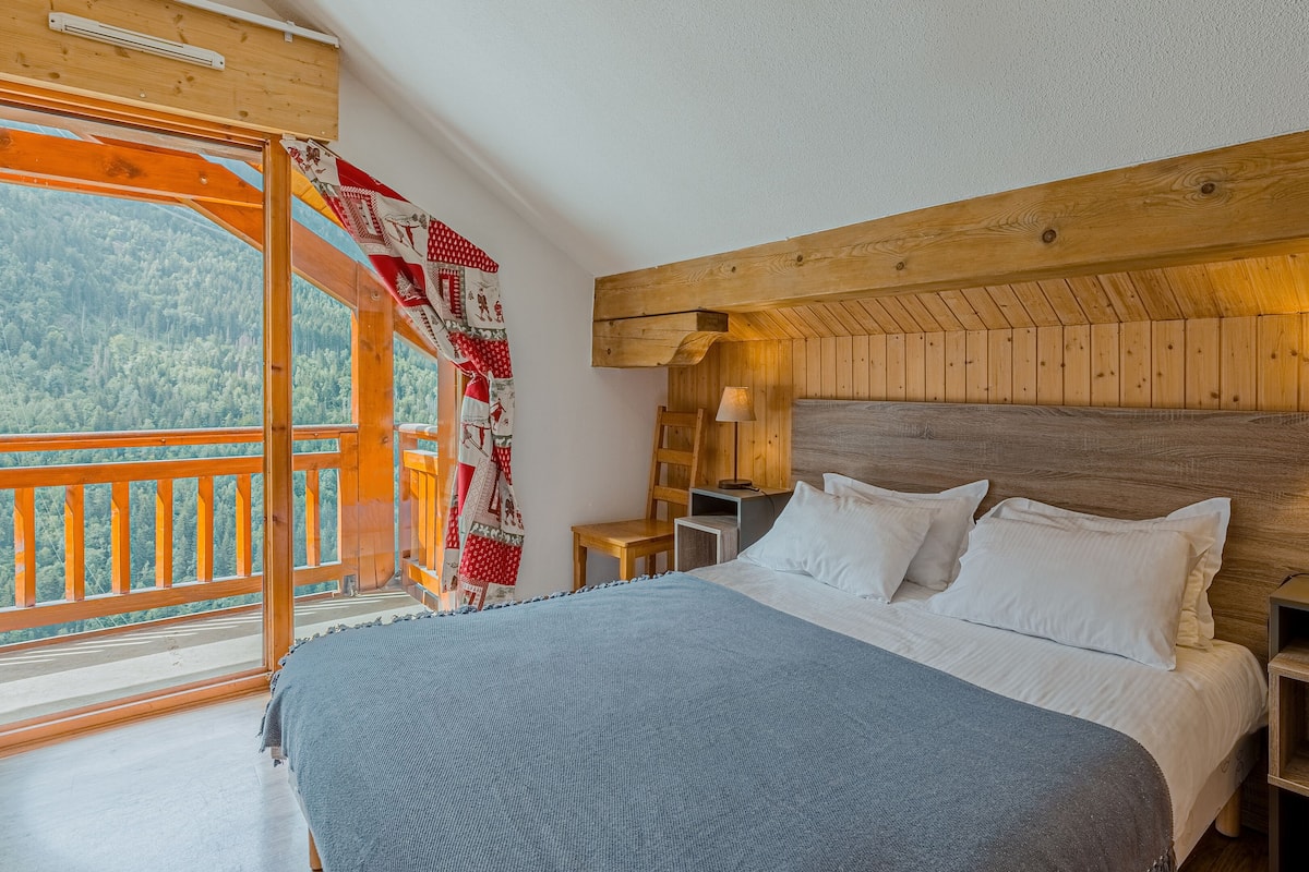 Quarto com cabeceira de madeira, cama com roupa de cama branca, mesa de cabeceira com luminária e vista para uma floresta através de uma grande janela.