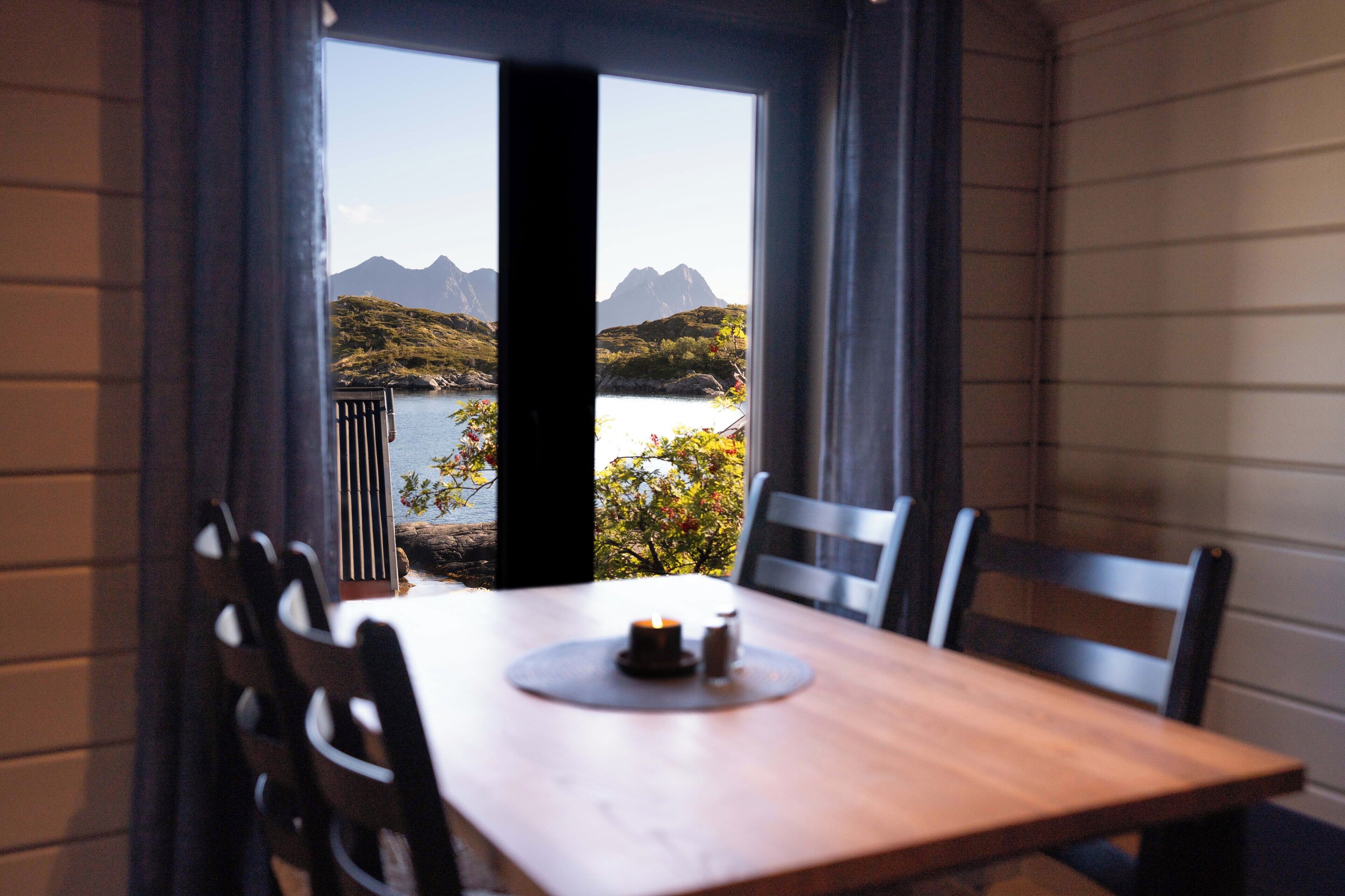 Uma mesa de jantar com cadeiras, uma vela e vista para um lago e montanhas através de uma janela.