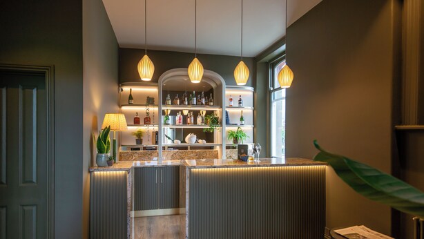 Uma área de bar moderna com um balcão central, prateleiras com garrafas e luminárias pendentes.