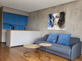 Uma sala de estar moderna com um sofá cinza, duas mesinhas redondas, uma máquina de café e uma área de cozinha com armários azuis.
