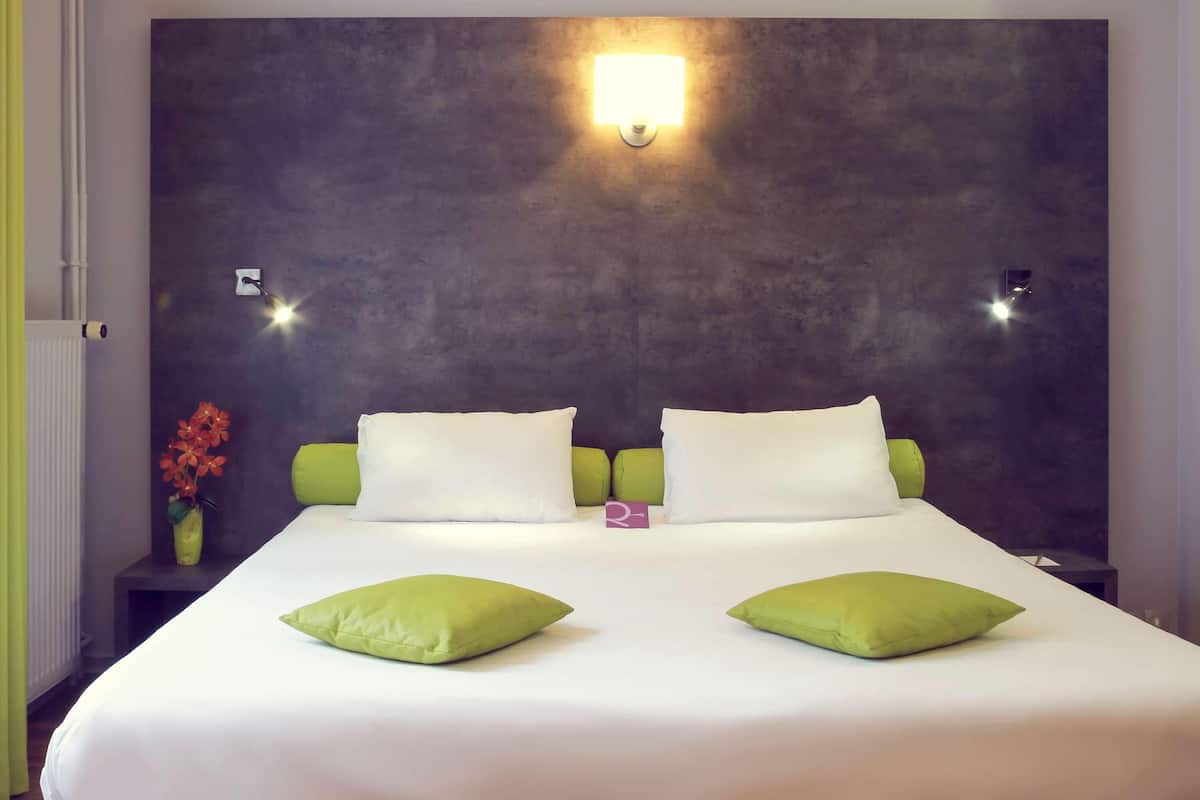Quarto de hotel com uma cama grande, duas almofadas, uma mesinha de cabeceira, um vaso com flores e um candeeiro fixado na parede.