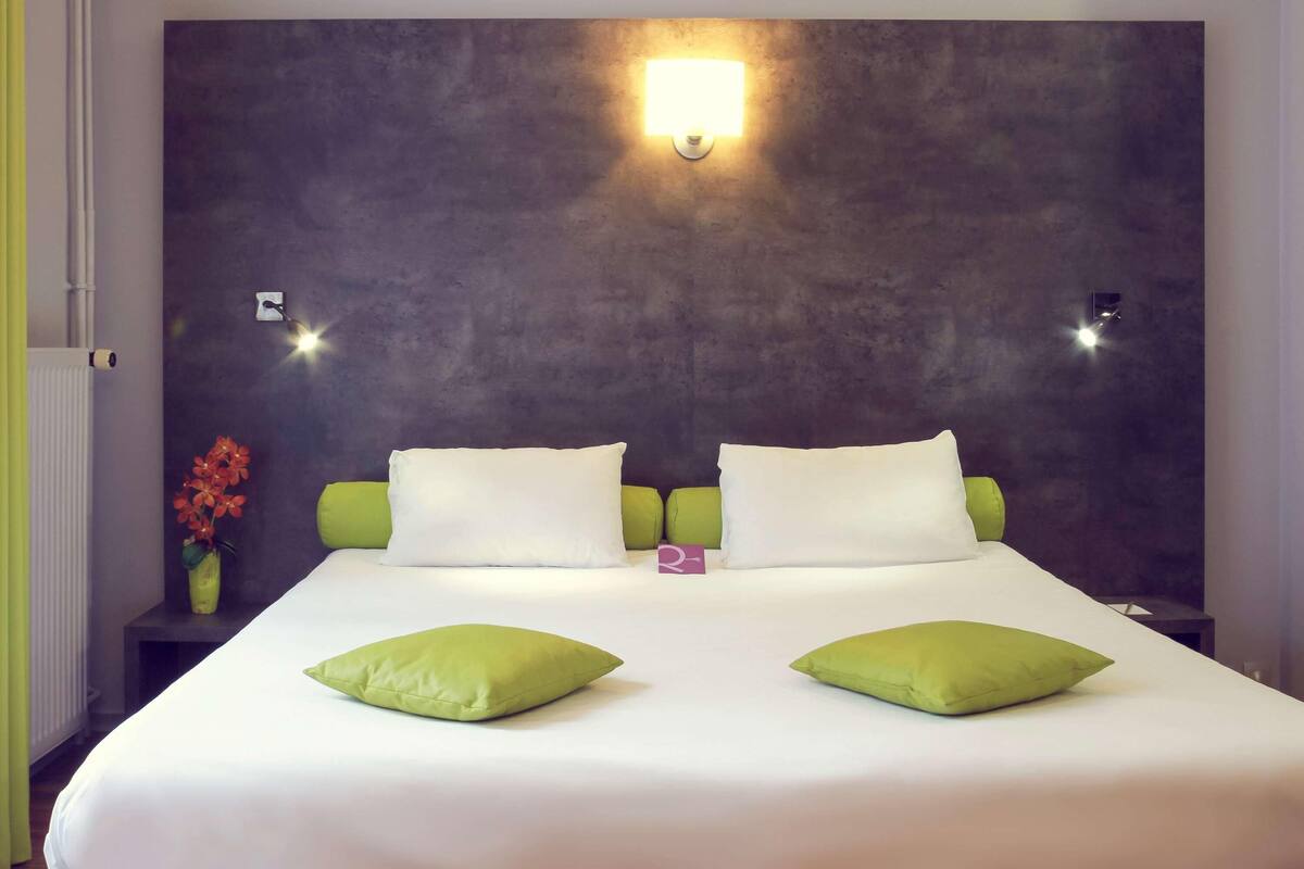 Quarto de hotel com uma cama grande, duas almofadas, uma mesinha de cabeceira, um vaso com flores e um candeeiro fixado na parede.