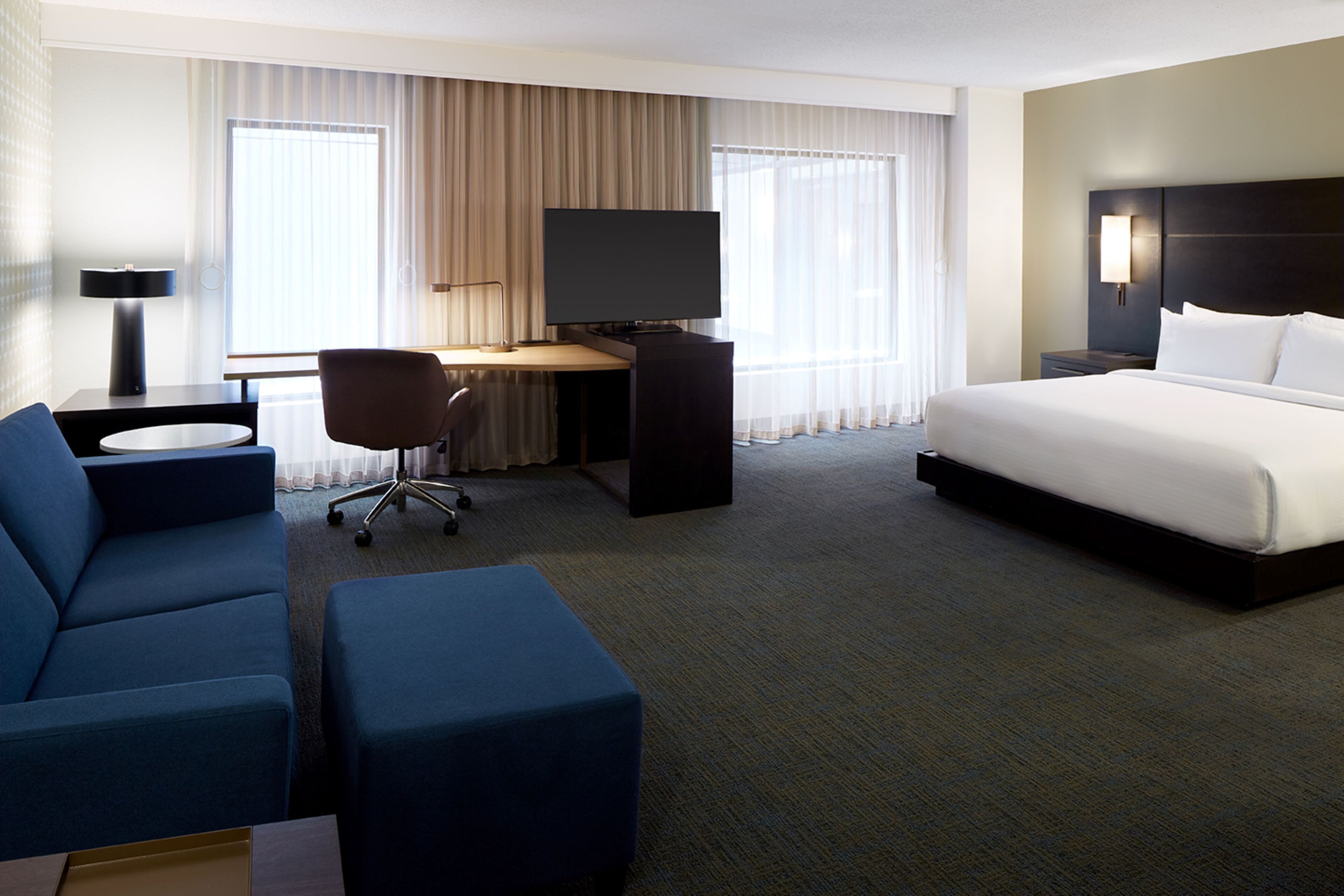 Quarto de hotel com cama, escrivaninha com computador, cadeira e sofá.
