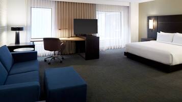 Quarto de hotel com cama, escrivaninha com computador, cadeira e sofá.