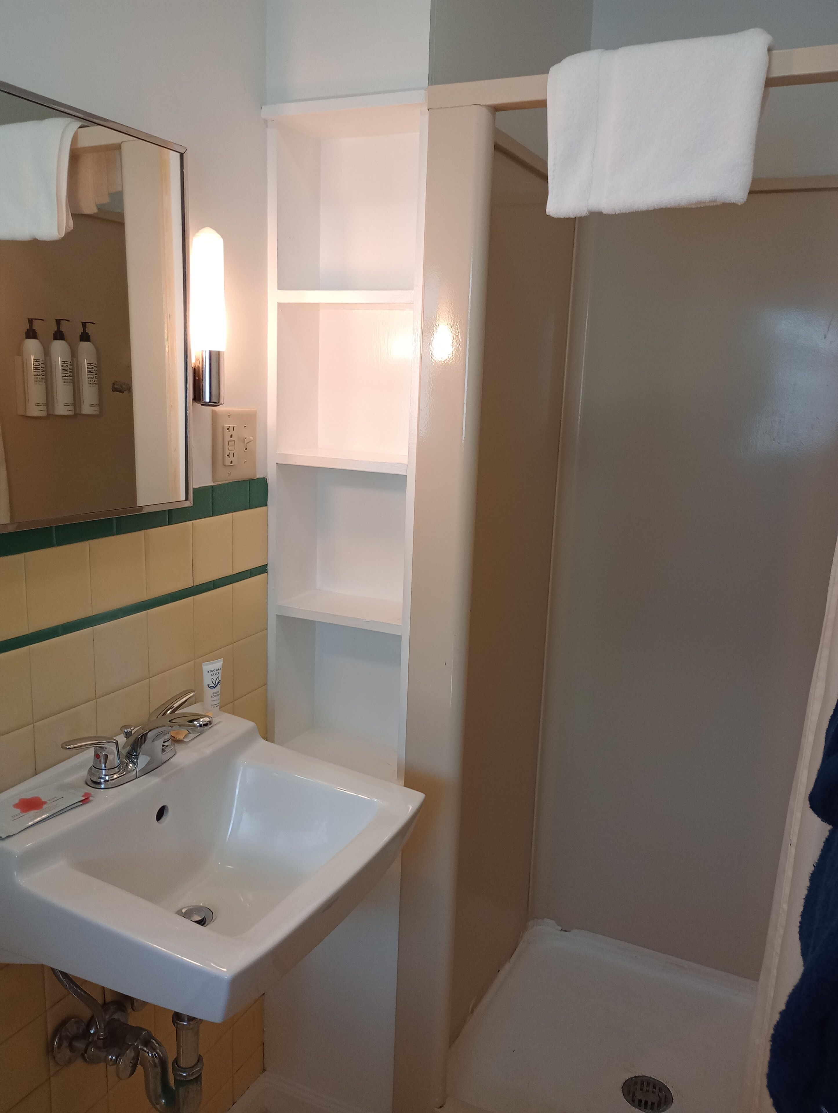 Casa de banho com lavatório, espelho e chuveiro. Há produtos de higiene em cima da parede e uma toalha pendurada na porta do chuveiro.