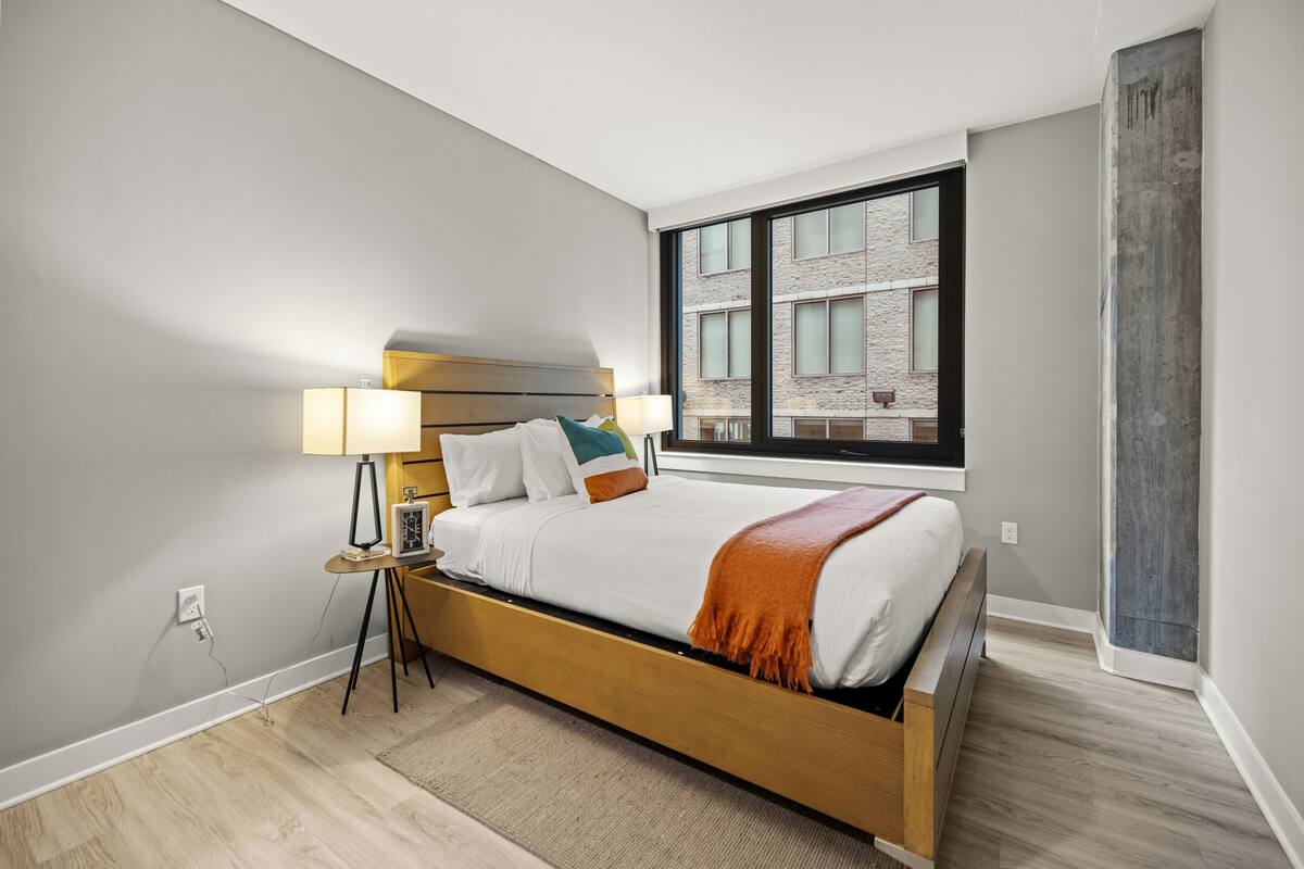Quarto de hotel moderno com cama de madeira, mesa de cabeceira com abajur, janela grande com vista para um edifício e piso claro.