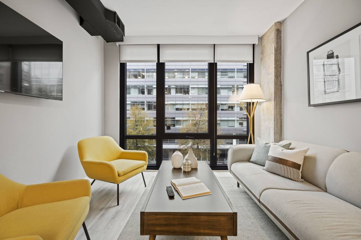 Uma sala de estar moderna com sofá, poltrona amarela e mesa de centro com controle remoto e um livro.