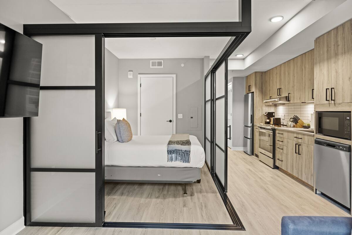 Quarto de hotel moderno com cama, mesa de cabeceira e vista para a área da cozinha.
