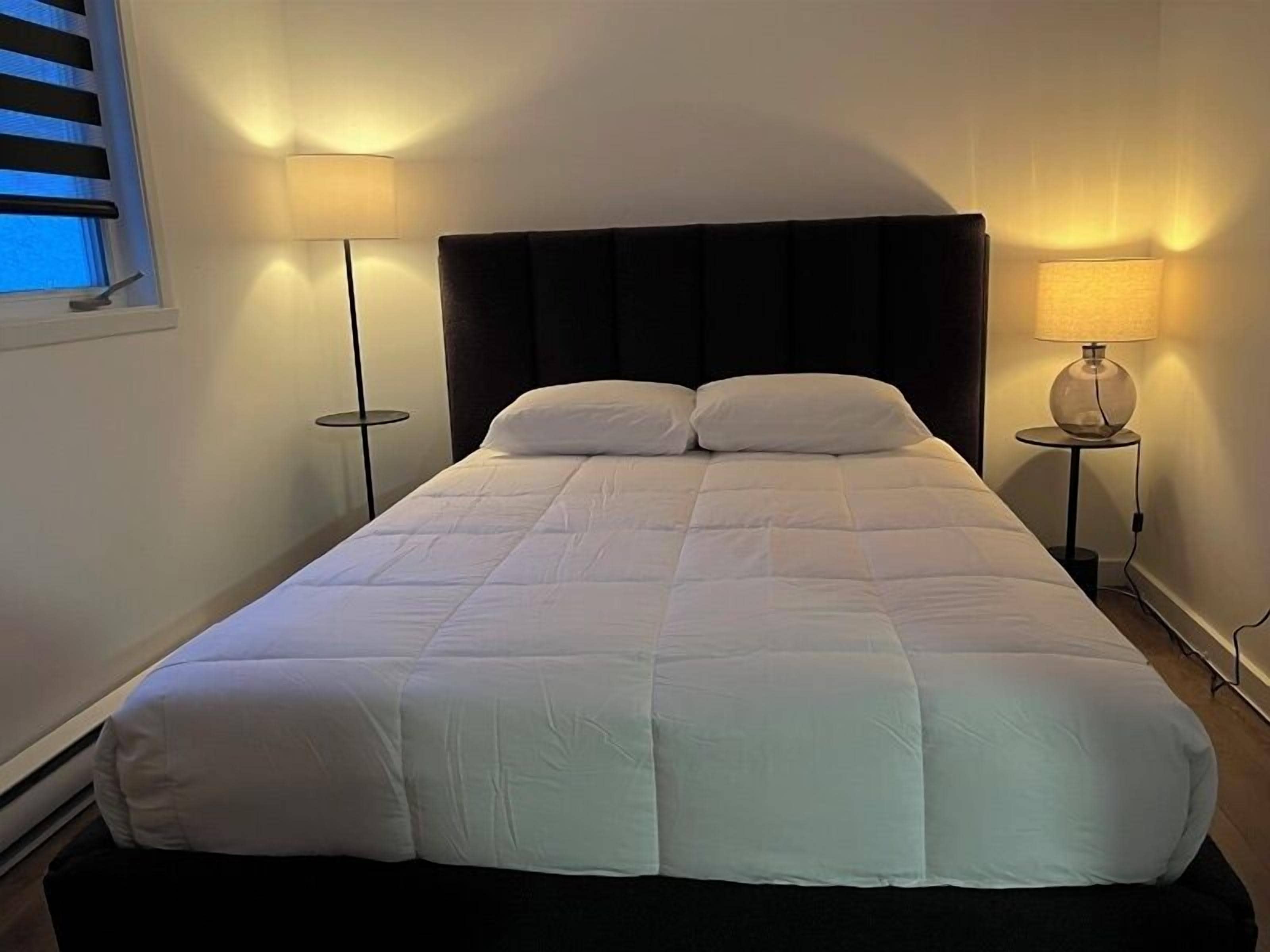 Uma cama bem arrumada, com cabeceira escura e duas luminárias laterais.