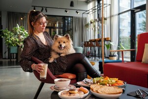 Uma mulher sentada em uma cadeira com um cachorro, segurando uma bebida, e uma mesa com comida e uma tigela em um ambiente interno moderno.