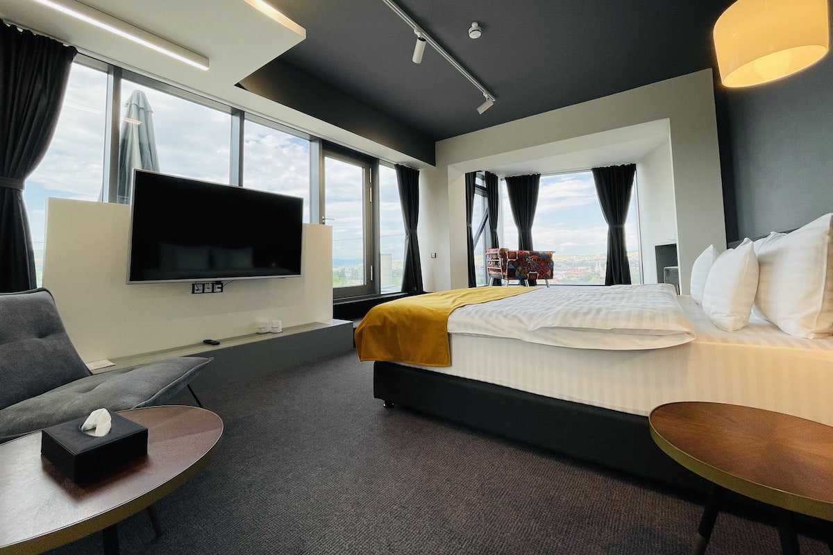 Quarto de hotel moderno com uma cama grande, televisão de tela plana, sofá e uma mesa pequena.