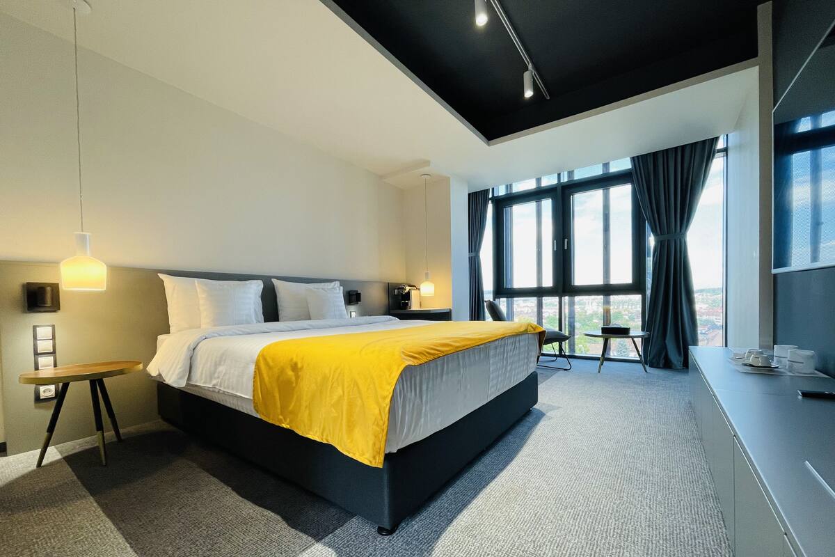 Quarto de hotel moderno com uma cama grande, uma escrivaninha e vista da cidade através de amplas janelas.
