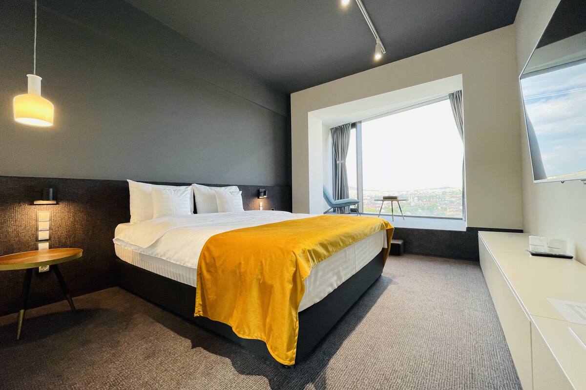 Quarto de hotel moderno com uma cama grande, uma escrivaninha e vista da cidade através de uma janela ampla.